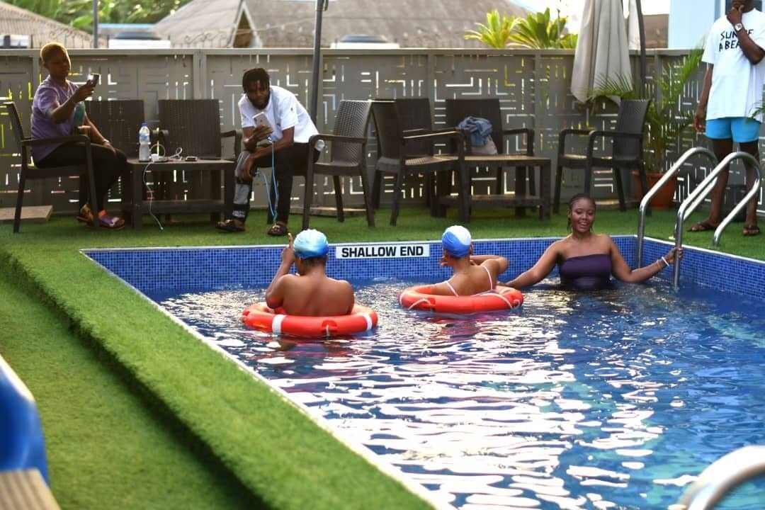 Piscina com duas pessoas na água, uma área rasa e um homem sentado numa cadeira usando um celular.