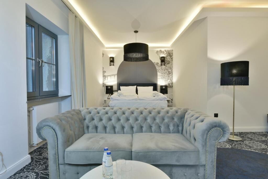 Quarto de hotel com sofá cinza estofado, mesa de centro redonda, cama com roupa de cama branca, luminária pendente preta e tapete estampado.