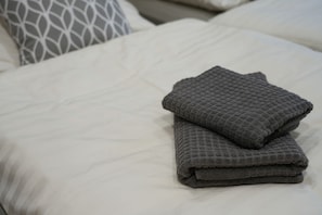 Cama bem arrumada com lençol branco e duas mantas cinzas dobradas.