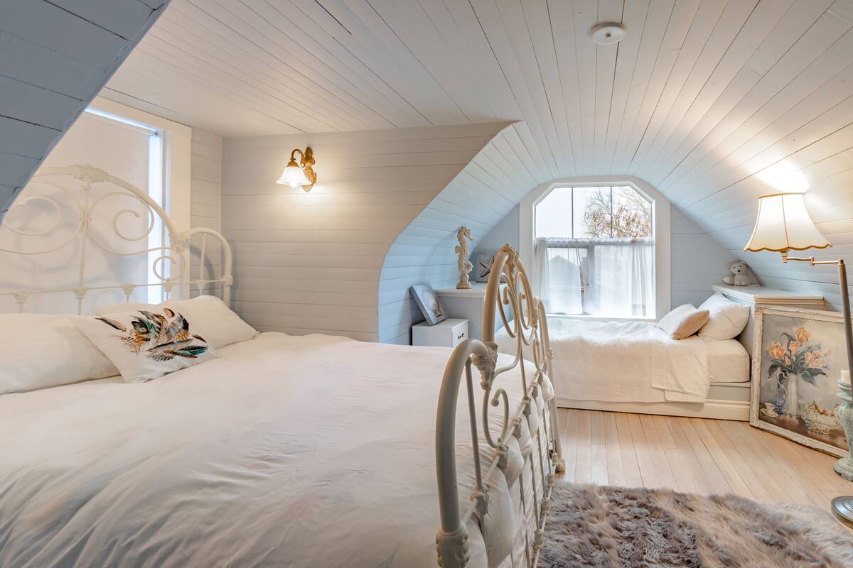 Quarto com teto inclinado, cama com roupa de cama branca, mesa de cabeceira, abajur e um quadro emoldurado.