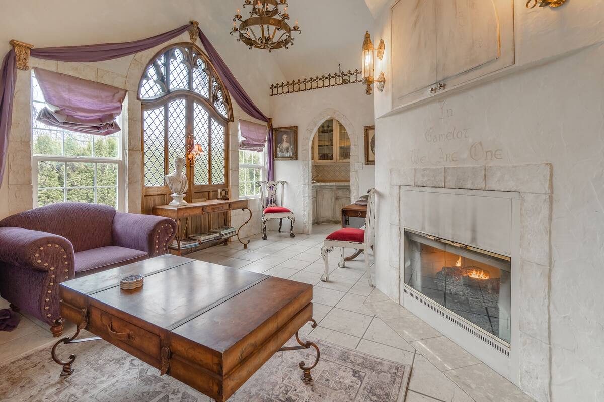 Quarto com lareira, uma mesa de madeira, um sofá roxo e uma janela com vista para o verde.