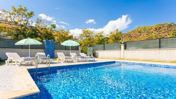 Piscina com espreguiçadeiras e guarda-sóis, cercada por um muro de tijolos e árvores.