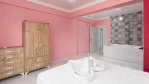 Quarto com paredes cor-de-rosa, um guarda-roupa de madeira, uma cama branca com roupão branco, uma toucador branca com espelho e uma área de chuveiro.