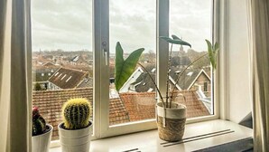 Janela com vista para telhados e paisagem urbana. Há três vasos de plantas no parapeito.