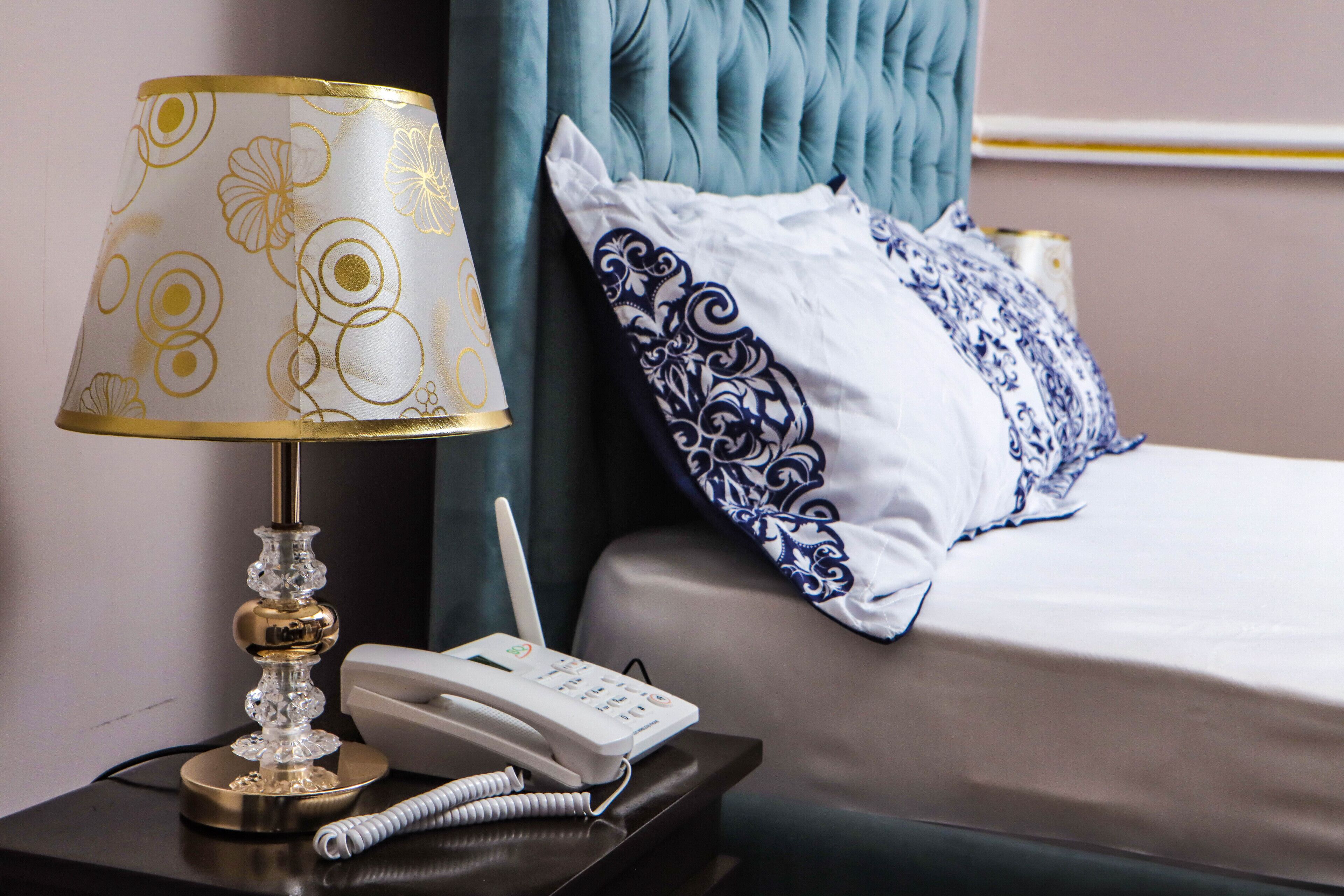 Quarto de hotel com mesa de cabeceira que inclui uma luminária, telefone e um objeto decorativo. A cama possui cabeceira acolchoada e almofadas estampadas em azul e branco.