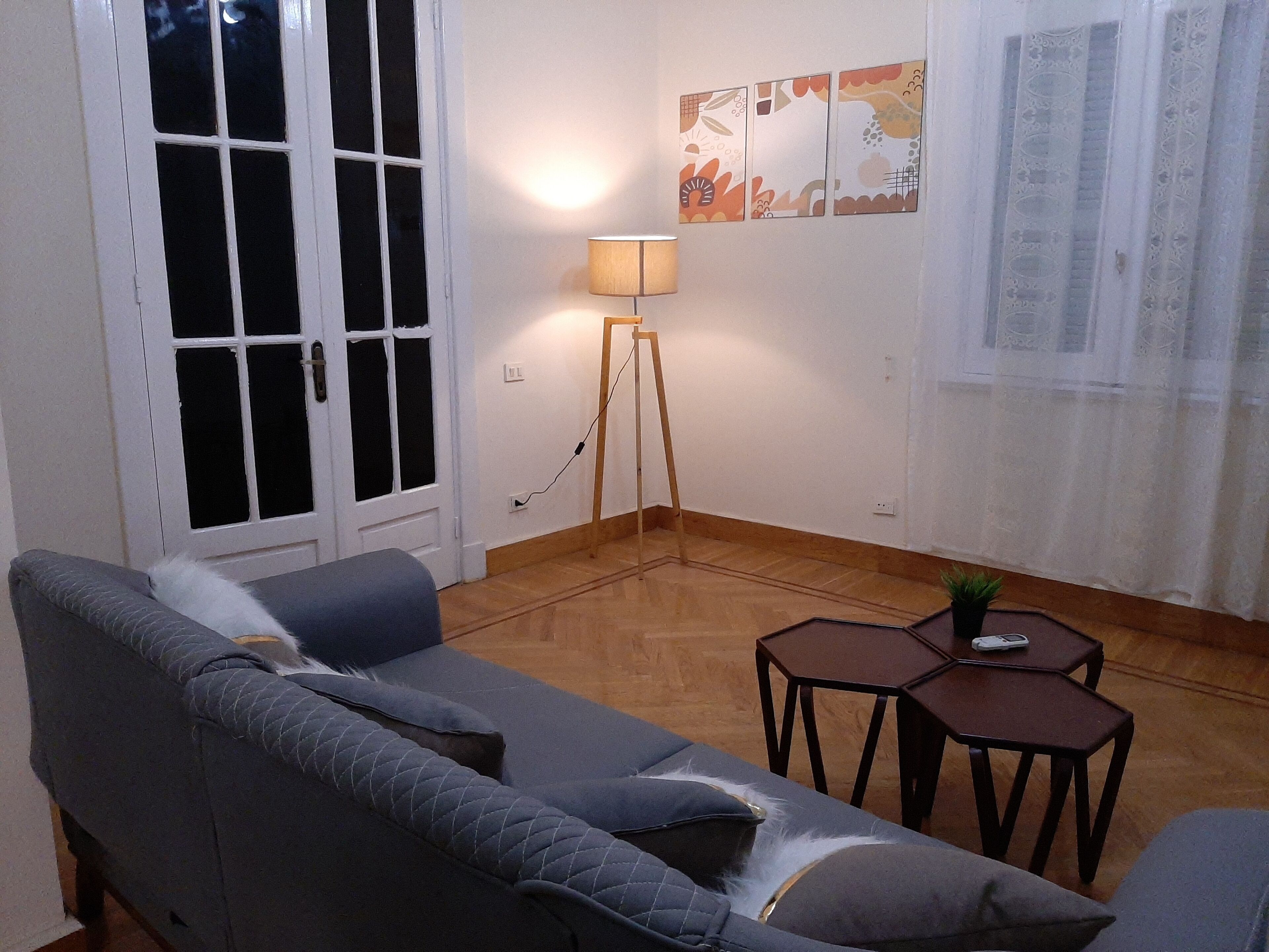 Uma sala de estar com um sofá cinzento, uma mesa pequena, uma luminária e uma janela com cortinas.