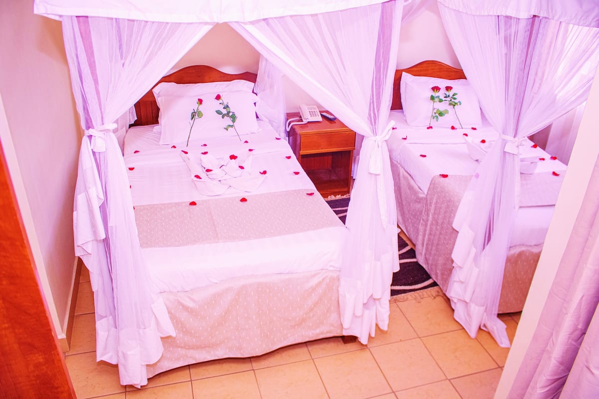 Quarto com duas camas cobertas por lençóis brancos, cada uma com dossel. Um criado-mudo de madeira fica entre as camas, com um telefone em cima.
