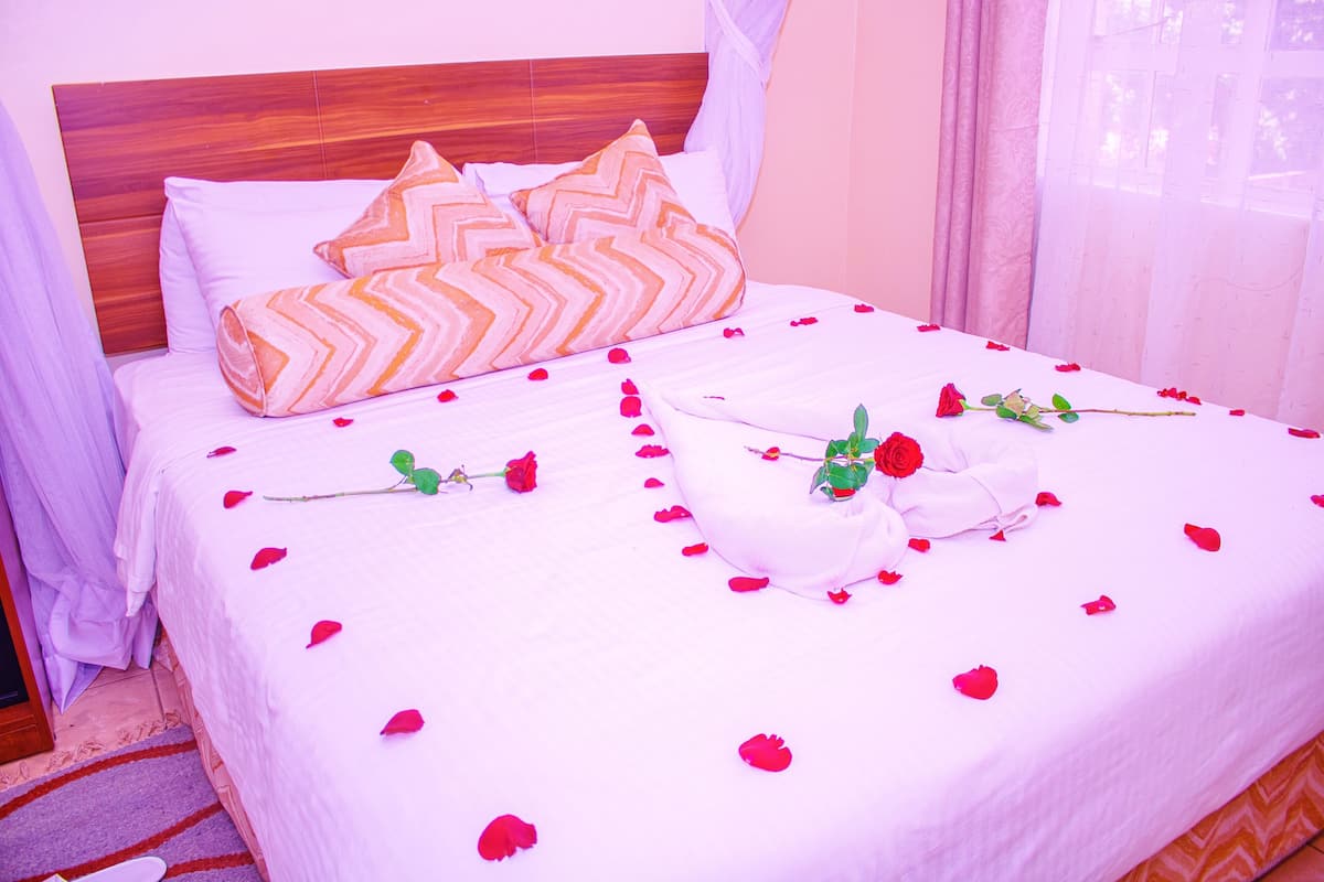 Cama bem arrumada com roupa de cama branca, várias almofadas e pétalas de rosa espalhadas pela superfície.