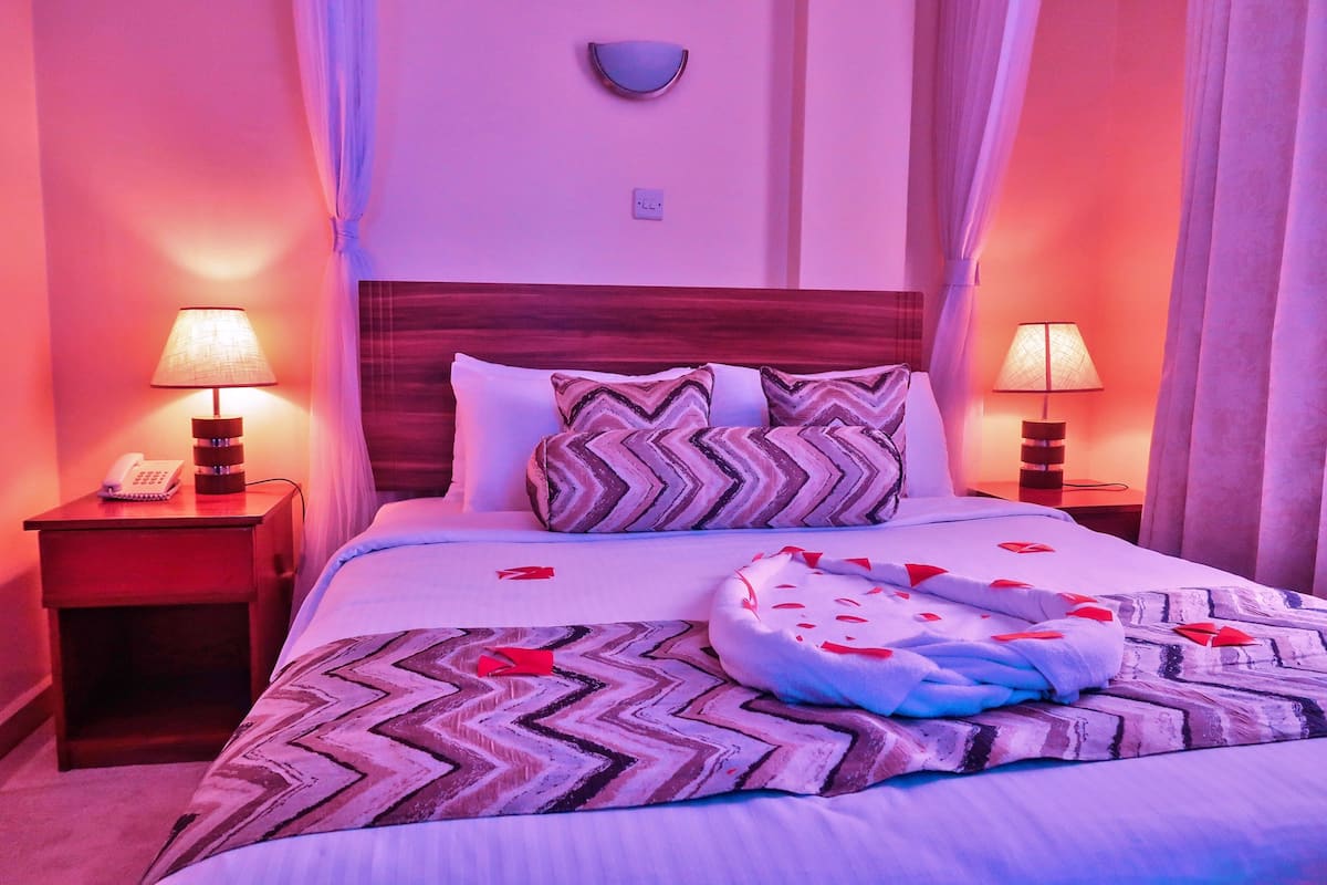 Quarto de hotel com cabeceira de madeira, duas mesas de cabeceira com luminárias, cama com almofadas estampadas e telefone ao lado da cama.
