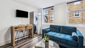 Uma sala de estar moderna com um sofá azul, uma mesa de jantar de madeira com duas cadeiras, uma TV de tela plana fixada na parede e grandes janelas com vista para edifícios de tijolo.