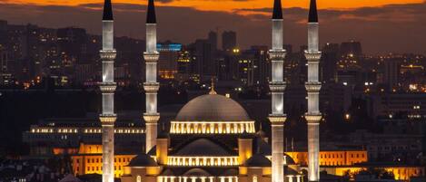 Uma mesquita com quatro minaretes e uma cúpula grande, cercada por um horizonte urbano ao anoitecer.