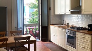 Cozinha compacta com armários brancos, fogão a gás e forno embutido. Uma área de refeições com mesa e cadeiras é visível, e uma porta de vidro deslizante dá acesso a uma área externa com plantas.