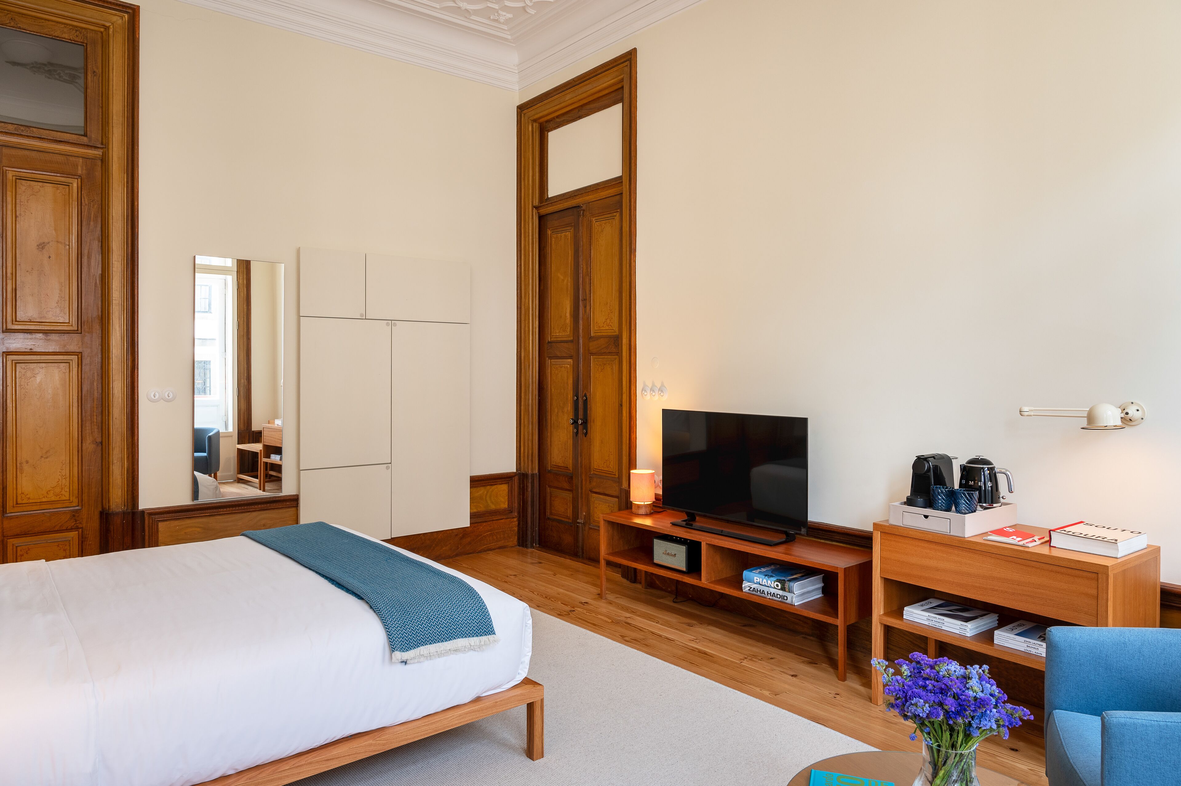 Quarto de hotel com cama, televisão, escrivaninha com abajur, cafeteira e um vaso com flores.