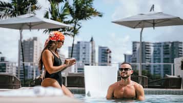 Um homem e uma mulher em uma piscina, com guarda-sóis e palmeiras ao fundo.