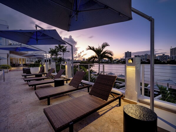 Um lounge no terraço com espreguiçadeiras, guarda-sóis e vista para o horizonte da cidade ao entardecer.