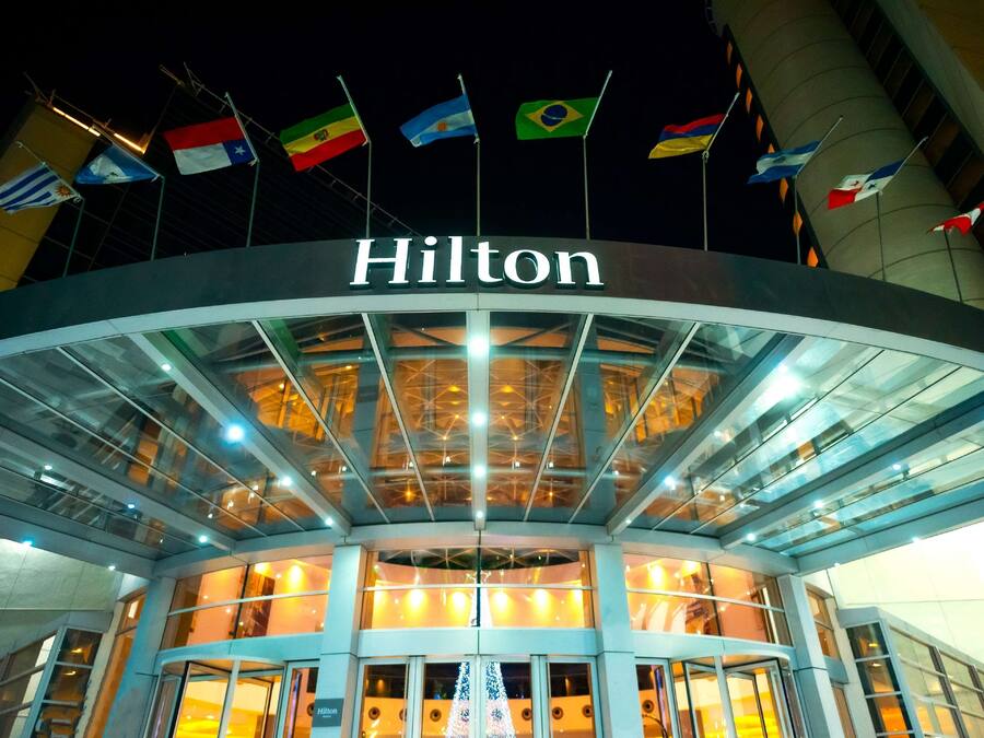 Entrada de um hotel Hilton, com várias bandeiras, um toldo de vidro e interior bem iluminado.