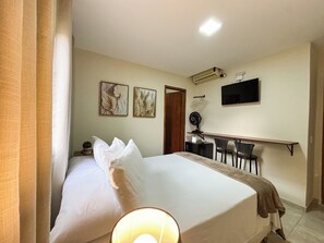 Quarto de hotel com cama, escrivaninha com cadeiras, televisão e duas obras de arte penduradas na parede.