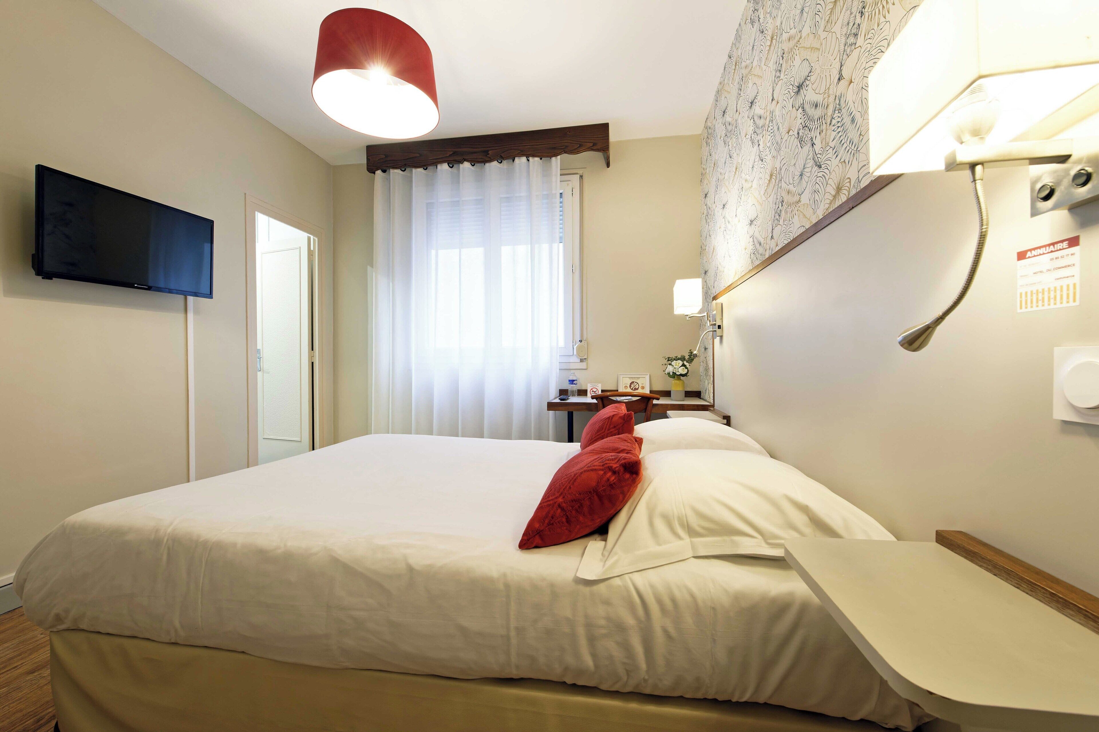 Quarto de hotel com uma cama grande, uma televisão de tela plana na parede, uma escrivaninha com uma cadeira e uma janela com cortinas translúcidas.