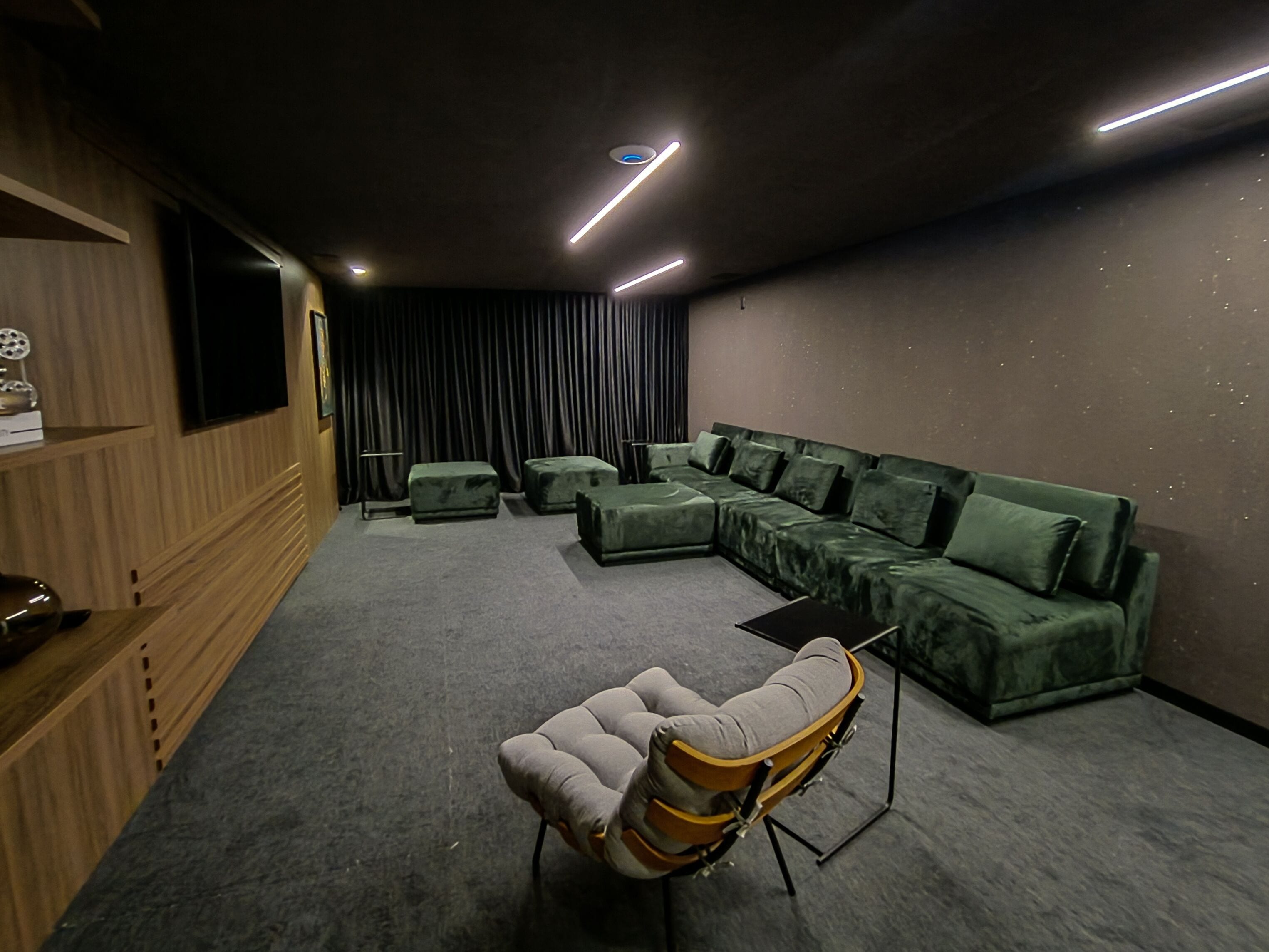 Uma sala de cinema moderna com um sofá seccional, uma poltrona confortável e uma TV de tela plana em frente a um fundo de cortina escura.