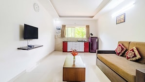 Uma sala de estar com televisão, uma mesa de centro com flores, um sofá com almofadas decorativas e uma cozinha ao fundo.