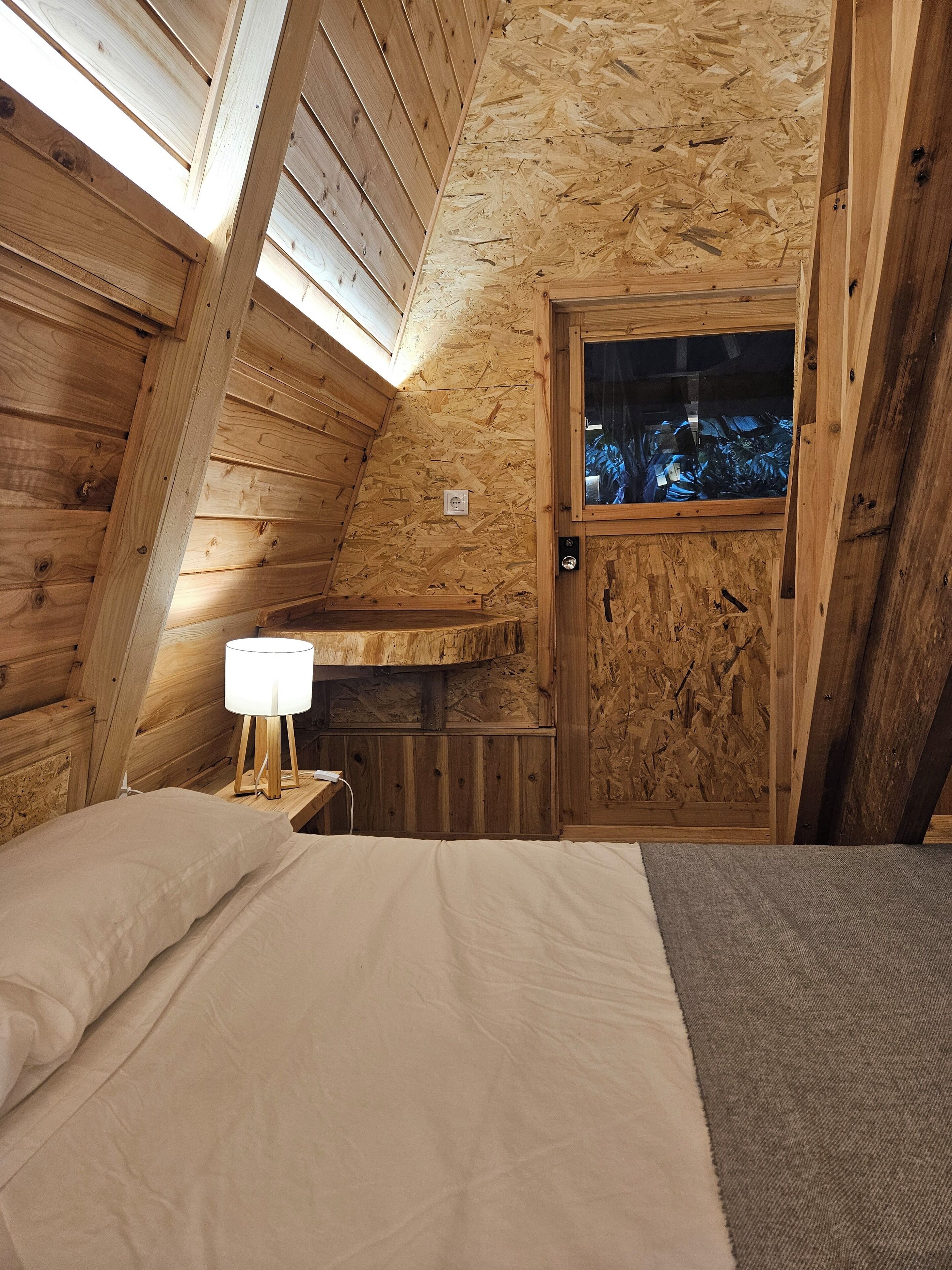 Um quarto de cabana de madeira aconchegante com teto inclinado, uma cama com lençóis brancos, uma mesa de cabeceira com abajur e uma janela com vista para a floresta.