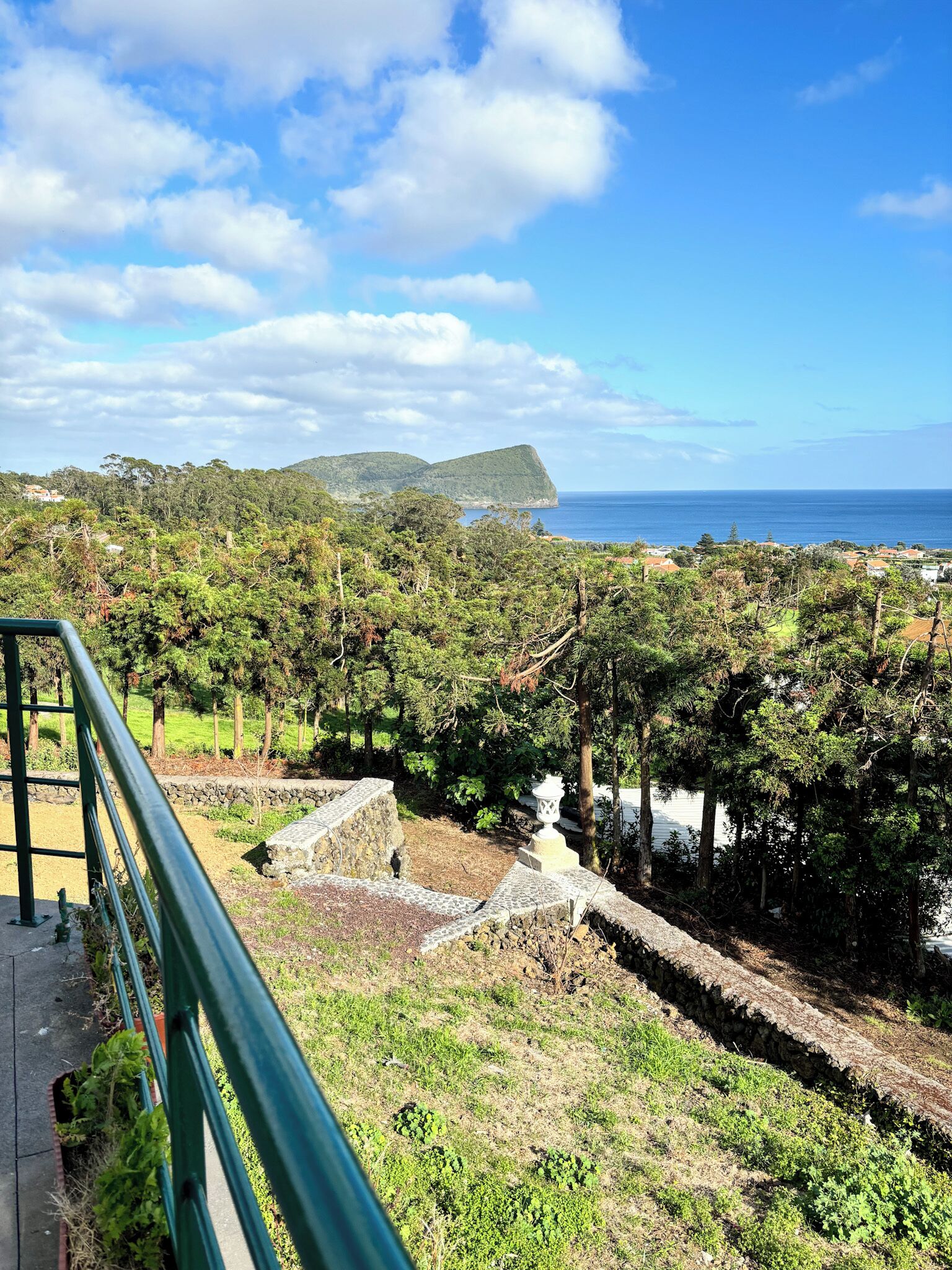 Paisagem costeira com um caminho de pedra, vegetação exuberante e vista para o mar e colinas ao longe.