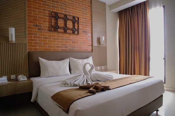 Quarto de hotel com parede de tijolos, cama com roupa de cama branca, manta marrom e arranjo de toalhas em formato de coração.