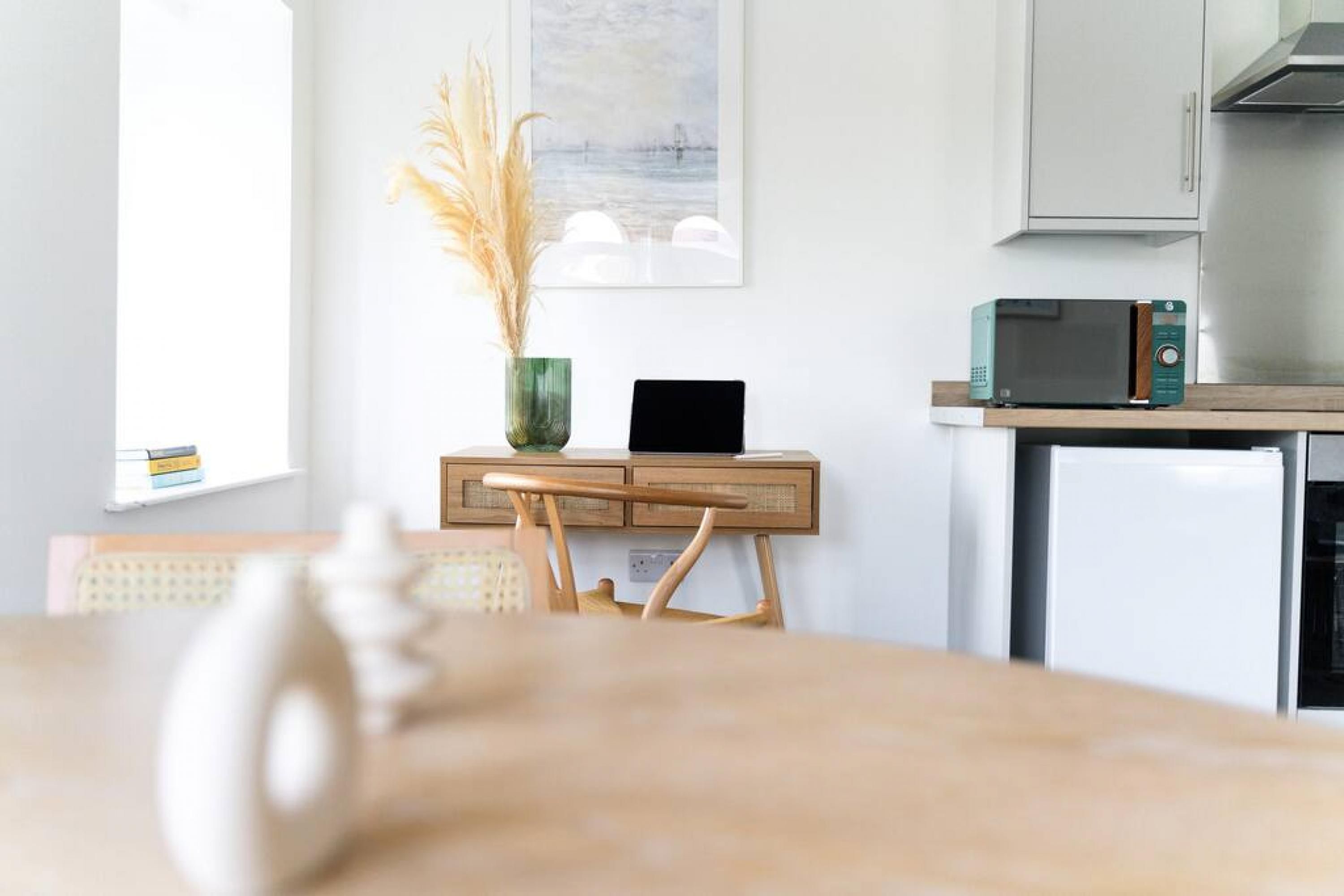 Uma cozinha com micro-ondas, mini geladeira, escrivaninha de madeira com laptop e um vaso com plantas secas.