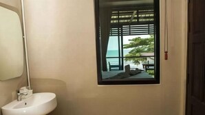 Casa de banho com uma pia branca, um espelho e uma janela com vista para o mar e palmeiras.