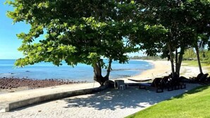 Uma praia com espreguiçadeiras e uma mesa pequena sob uma árvore, com vista para o oceano.