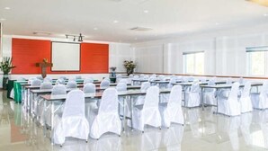 Sala de conferências com fileiras de mesas e cadeiras, uma tela de projeção e uma parede com detalhes em vermelho.