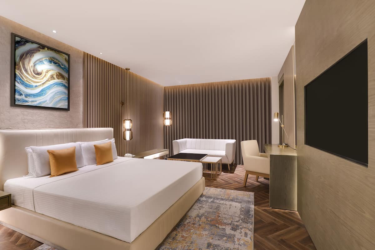 Quarto de hotel moderno com uma cama grande, um sofá, uma TV e um quadro grande na parede.