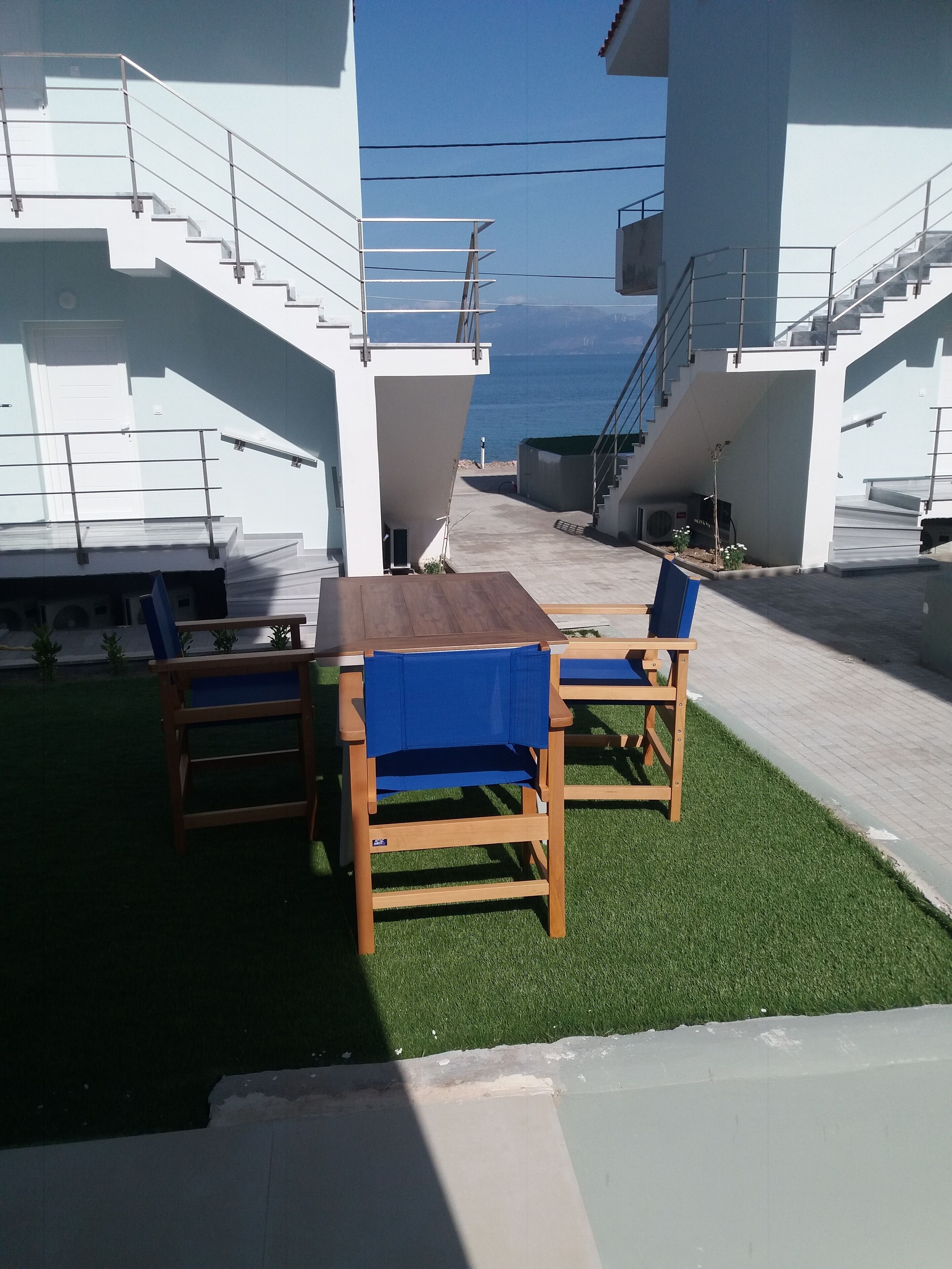 Uma área de estar moderna ao ar livre, com mesa de madeira e duas cadeiras azuis, cercada por grama artificial verde e paredes brancas.
