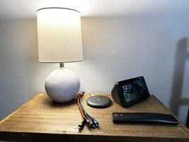 Uma mesa de cabeceira com uma lâmpada, um relógio digital, um controle remoto e um cabo de carregamento.