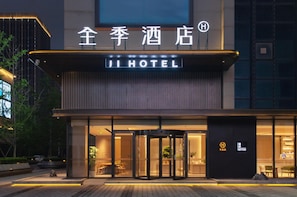 Entrada moderna de hotel com porta giratória, uma placa com a inscrição 'JI HOTEL' e um logotipo com a letra 'H' à direita.