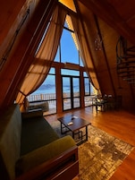 Um interior aconchegante, com um sofá, uma mesa e vista para o mar através de grandes janelas.