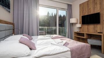 Quarto de hotel moderno com uma cama grande, uma escrivaninha, uma televisão e uma varanda com vista para árvores.