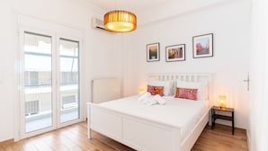 Um quarto com cama branca, mesa de cabeceira com abajur, e três quadros na parede.