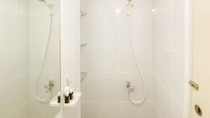 Casa de banho moderna com azulejos brancos, uma prateleira de vidro com produtos de higiene e um chuveiro com chuveiro manual.