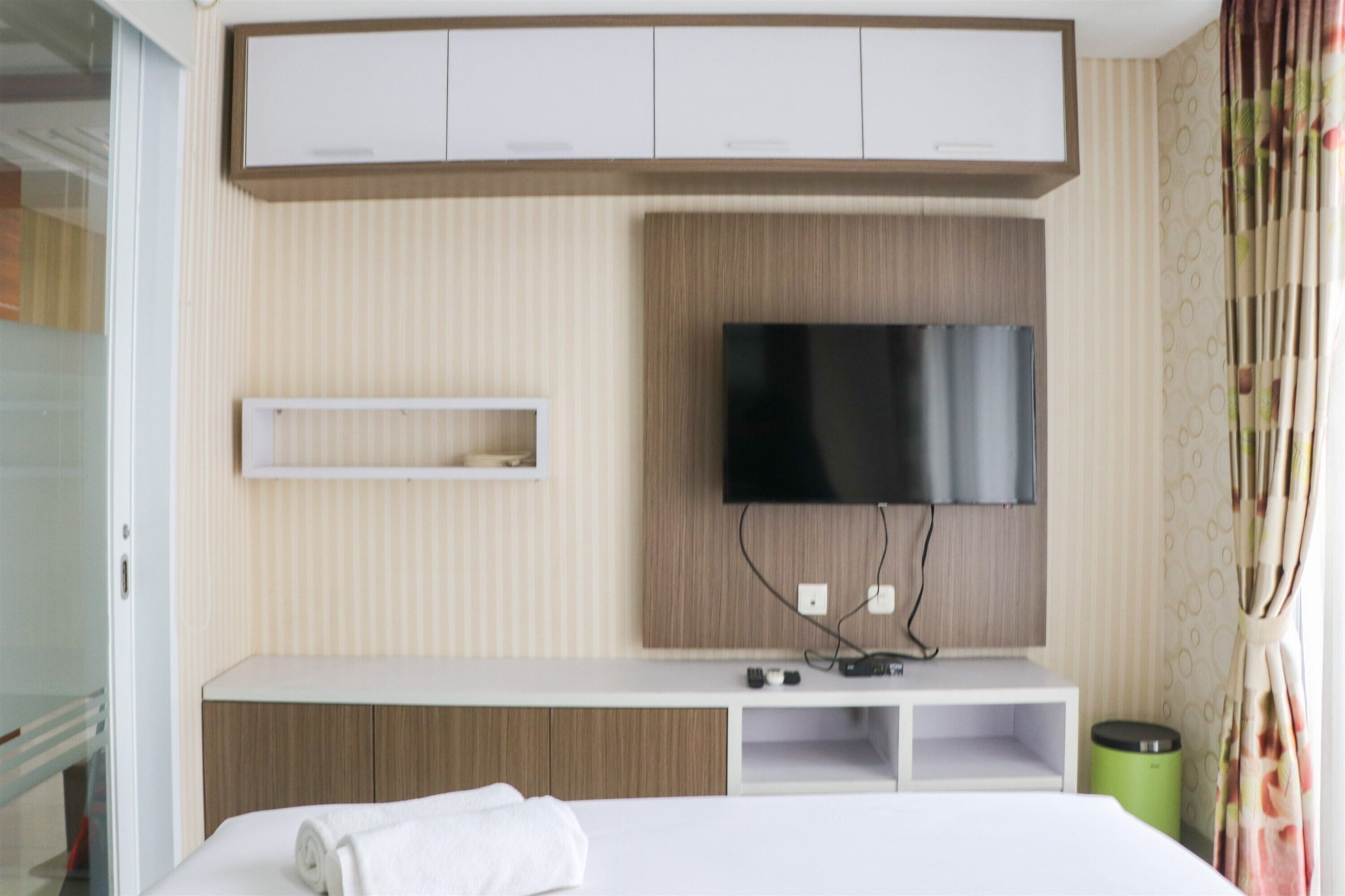 Quarto de hotel com uma cama, TV fixada na parede, prateleira pequena e lixeira.