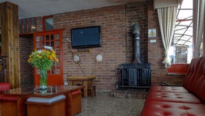 Uma sala de estar com parede de tijolos, uma televisão montada acima de uma lareira a lenha preta, uma mesa de madeira com um vaso de flores e um sofá de couro vermelho.