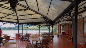 Uma área de refeições ao ar livre com mesas e cadeiras, vista para um lago e estrutura de cobertura.