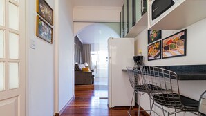 Um corredor estreito com piso de madeira, uma sala de jantar com cadeiras de metal e uma cozinha com armários e geladeira.