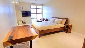 Quarto com cama de madeira, uma pequena mesa de cabeceira, uma cadeira, escrivaninha e televisão na parede.