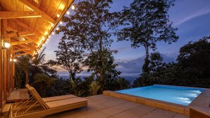 Piscina no terraço com deck de madeira e espreguiçadeiras, cercada por árvores e com vista para o céu.
