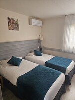 Quarto de hotel com duas camas, ar condicionado fixado na parede, abajur e janela com cortinas leves.