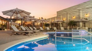 Piscina com espreguiçadeiras e guarda-sóis, rodeada por um edifício com fachada de vidro e iluminação externa.