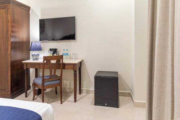 Quarto de hotel com uma escrivaninha de madeira, uma cadeira, televisão fixada na parede, um refrigerador preto pequeno e uma cama com colcha azul.
