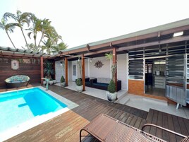 Quintal com piscina, deck de madeira e varanda coberta com assentos e plantas.