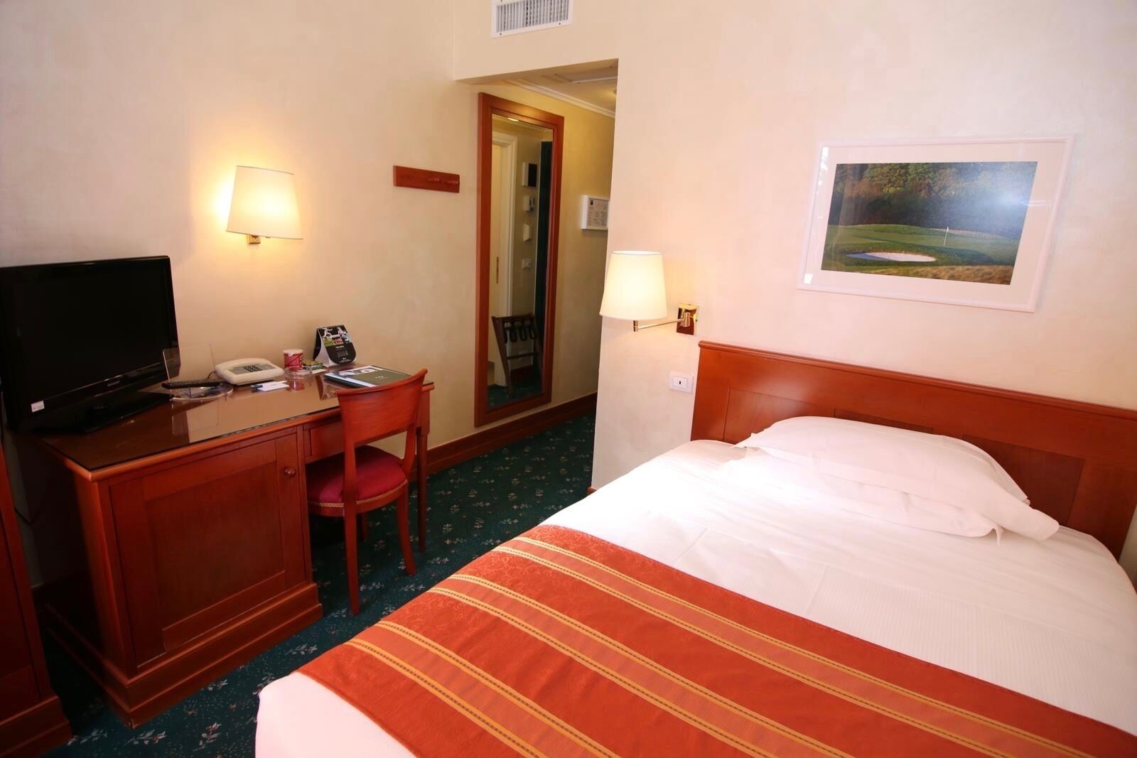 Quarto de hotel com cama, escrivaninha com televisão, cadeira, abajur e um quadro na parede.